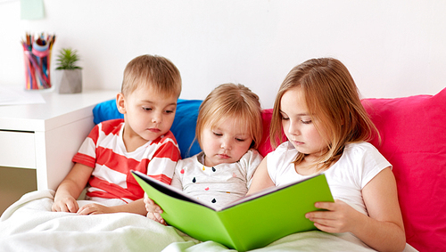 childhood, leisure and family concept - little kids reading book in bed ...