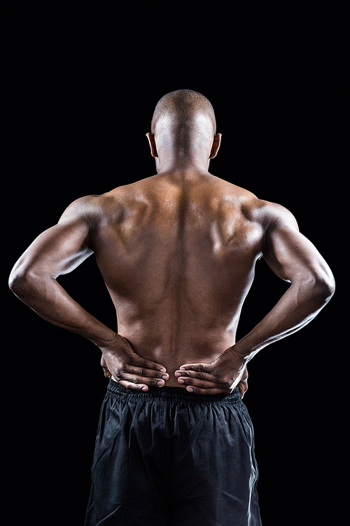 Rear view of muscular athlete stretching with hand on hip against black ...