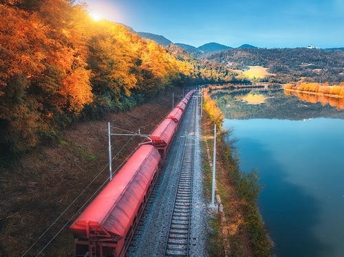 Aerial view of red freight train moving near river in alpine mountains ...