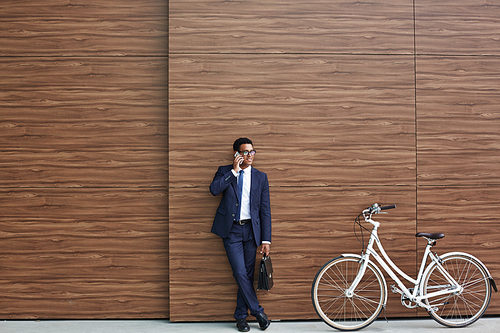 Modern agent in suit speaking on the phone by wooden wall with bicycle ...