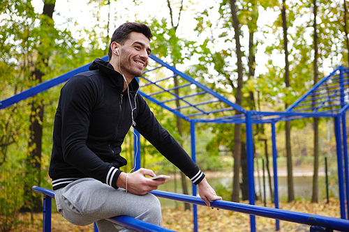 Cheerful young sporty man in headphones sitting on parallel bars in ...