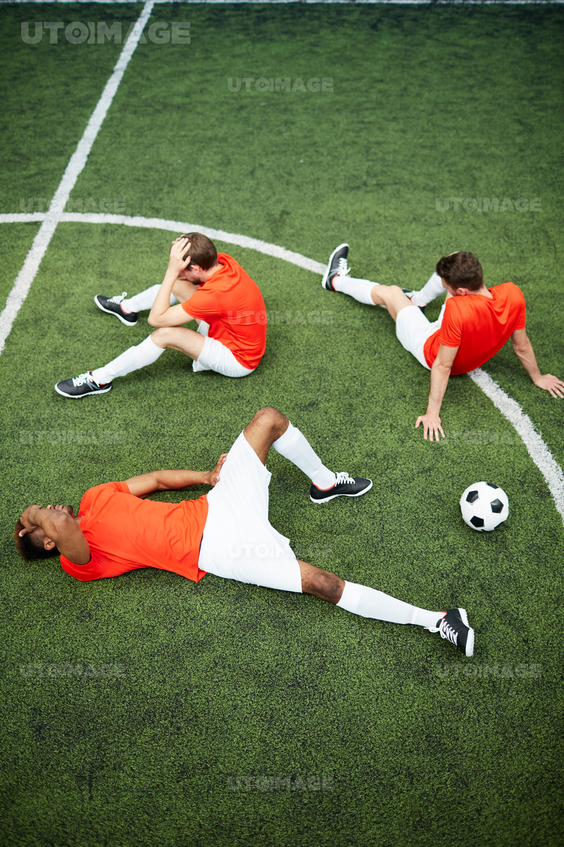 Three unlucky football players sitting and lying on green field after ...