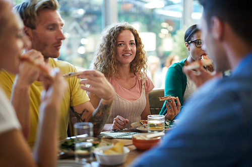 Casual girl with wavy hair eating pizza and looking at one of her ...