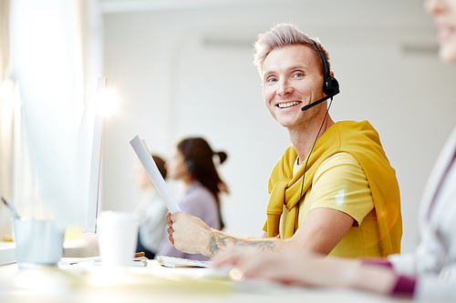 Young friendly opreator with headset by workplace among other staff ...