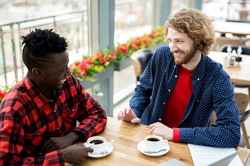Two friendly men in casualwear sitting by table in cafe and having ...