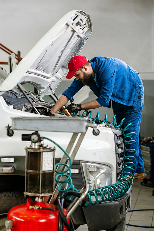 Young technician in workwear bending over open engine of large vehicle ...