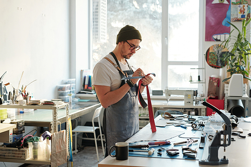 Young serious leatherworker in workwear standing by table in workshop ...