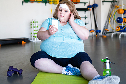 Full length portrait of obese young woman sitting on yoga mat on floor ...