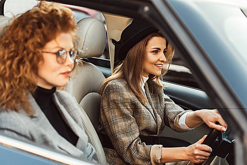 cheerful woman in black hat driving car while her ginger female friend ...