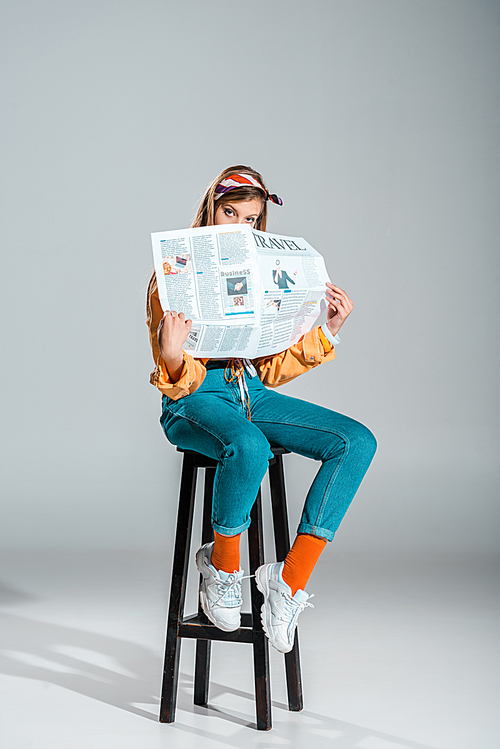young woman holding travel newspaper while sitting on stool on grey ...
