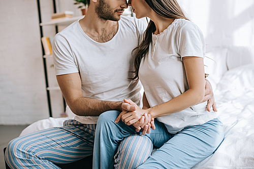 cropped shot of young couple cuddling while sitting on bed in morning ...