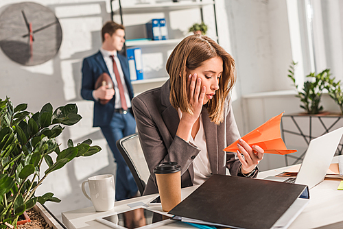 tired businesswoman holding paper plane near laptop with coworker on ...