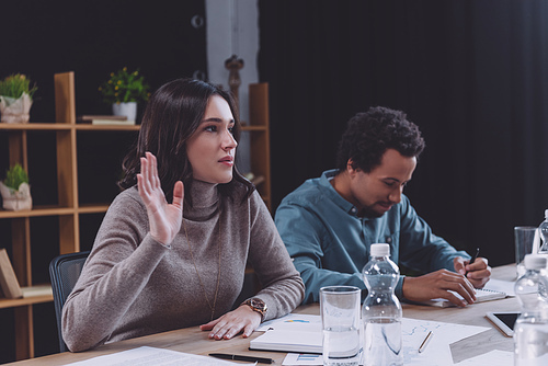 young businesswoman raising hand while sitting near african american ...