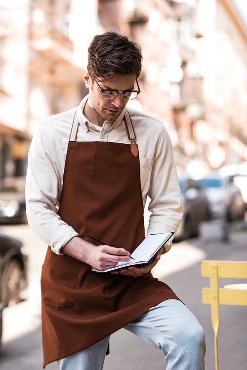 Concentrated barista in glasses and apron writing in notebook on street ...