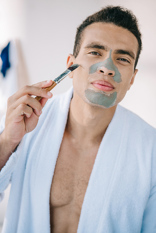 handsome man applying clay mask on face with brush while standing in ...