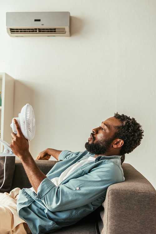 african american man holding blowing electric fan while lying on sofa ...