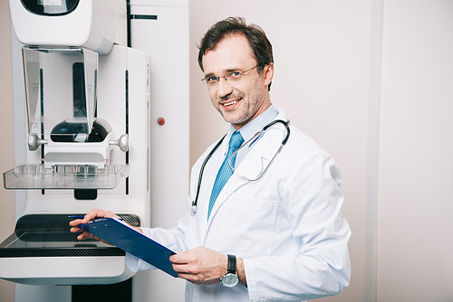 smiling radiologist holding clipboard while standing at x-ray machine ...