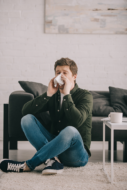 man sitting on carpet with crossed legs and sneezing in tissue near ...