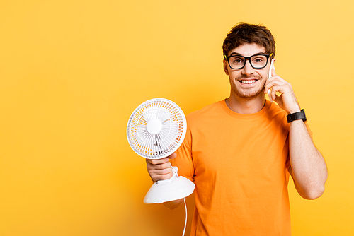 cheerful man talking on smartphone while holding electric fan on yellow ...