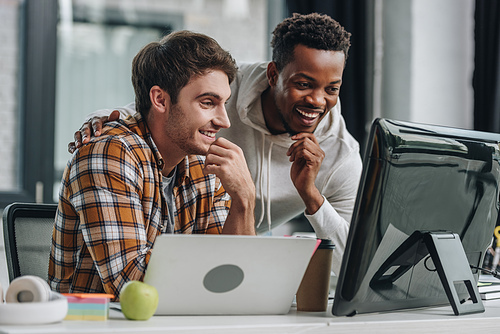 two cheerful multicultural programmers looking at computer monitor ...