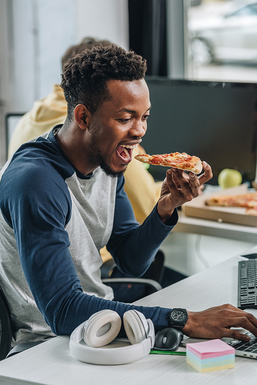 hungry african american programmer eating pizza while sitting at workplace near colleague | 유토 ...