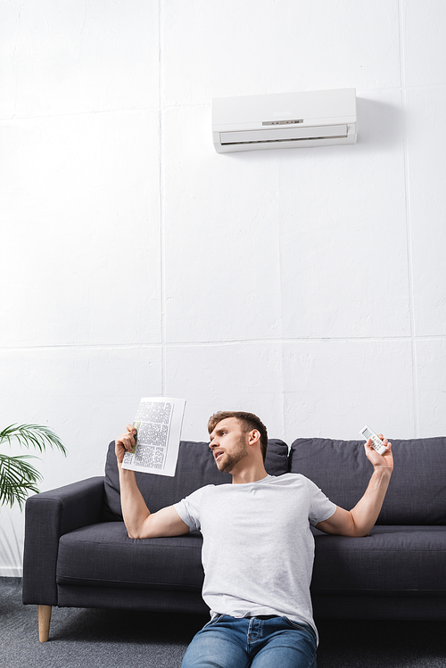 sad young man suffering from heat while using newspaper as hand fan at ...