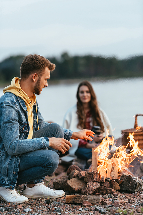 selective focus of handsome man sitting and looking at burning bonfire ...