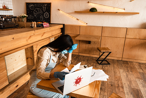 Stressed cafe owner in medical mask holding paper near laptop and ...